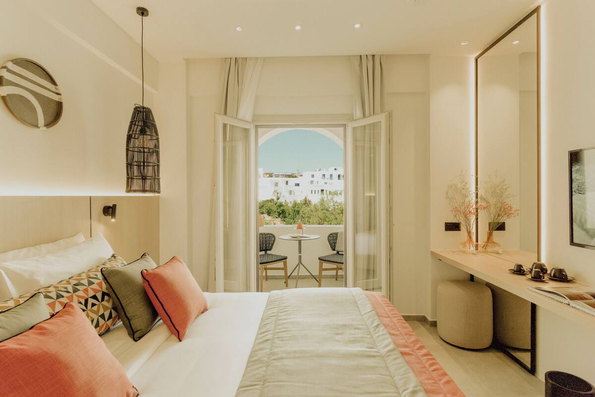 Quarto de hotel com cama, escrivaninha, cadeira e varanda com mesa e cadeiras.