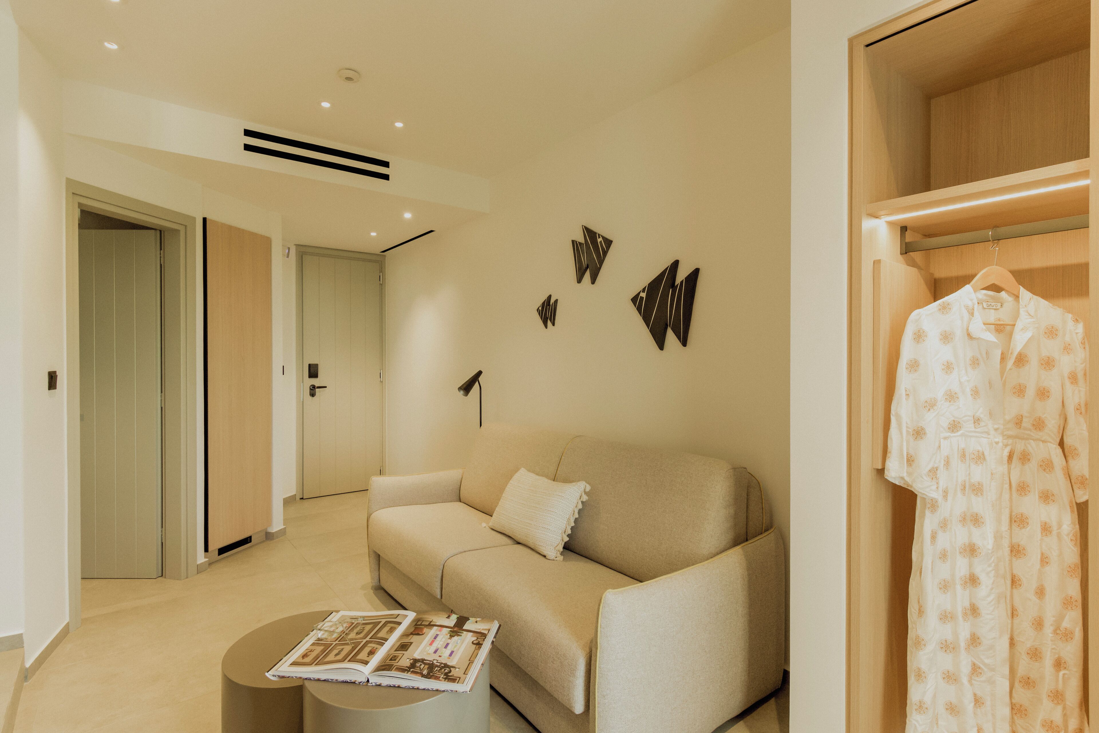 Uma sala de estar moderna com um sofá bege, uma mesa de centro redonda com uma revista e um guarda-roupa de madeira com cabides.