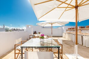 Terraço no telhado com mesa de vidro, cadeiras brancas e grandes guarda-sóis.