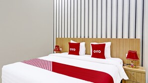 Quarto de hotel com cabeceira de madeira, cama com roupa de cama branca e vermelha, e mesa de cabeceira com abajur vermelho.