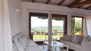 Uma sala de estar ampla com um sofá branco, uma mesa de centro e vista para um jardim e colinas através de grandes janelas.