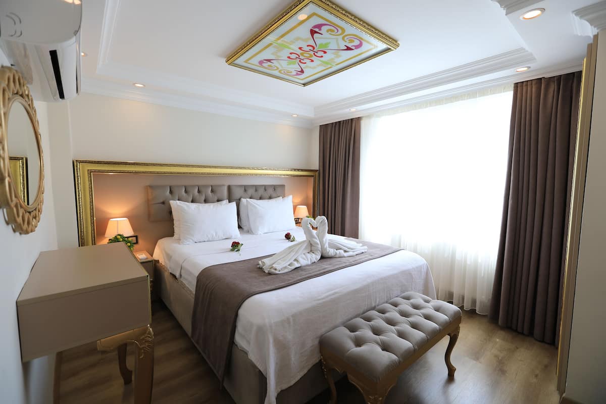 Quarto de hotel com uma cama grande, mesa de cabeceira, um banquinho e luminária no teto.