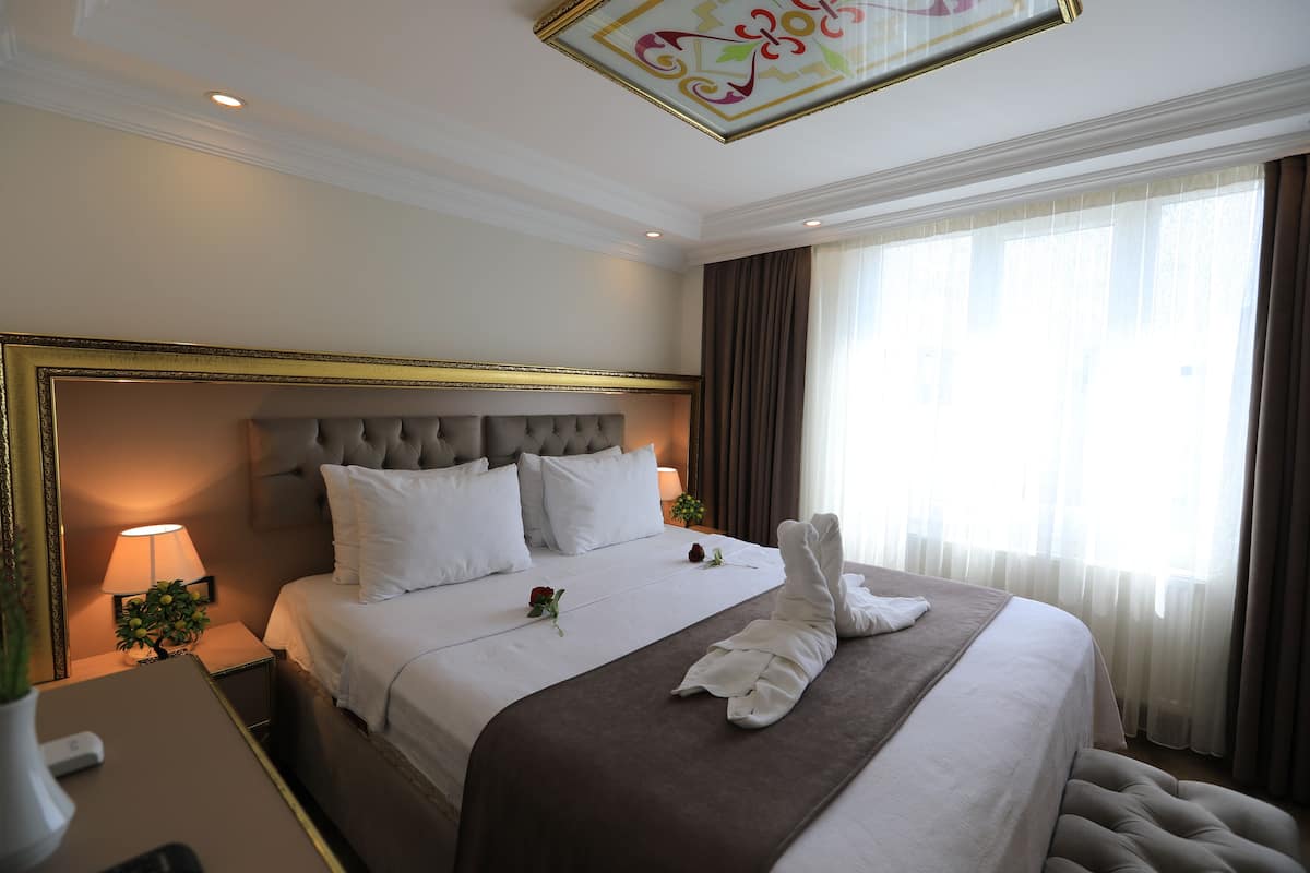 Cama bem arrumada com lençóis brancos e detalhes em castanho, uma mesa de cabeceira com abajur e uma pequena planta, e uma janela grande com cortinas.