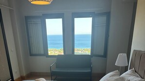 Quarto de hotel com cama, duas janelas vista para o mar, uma cadeira e um abajur.