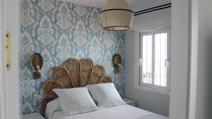 Quarto com papel de parede estampado, cabeceira de vime, roupa de cama branca e janela com persianas.