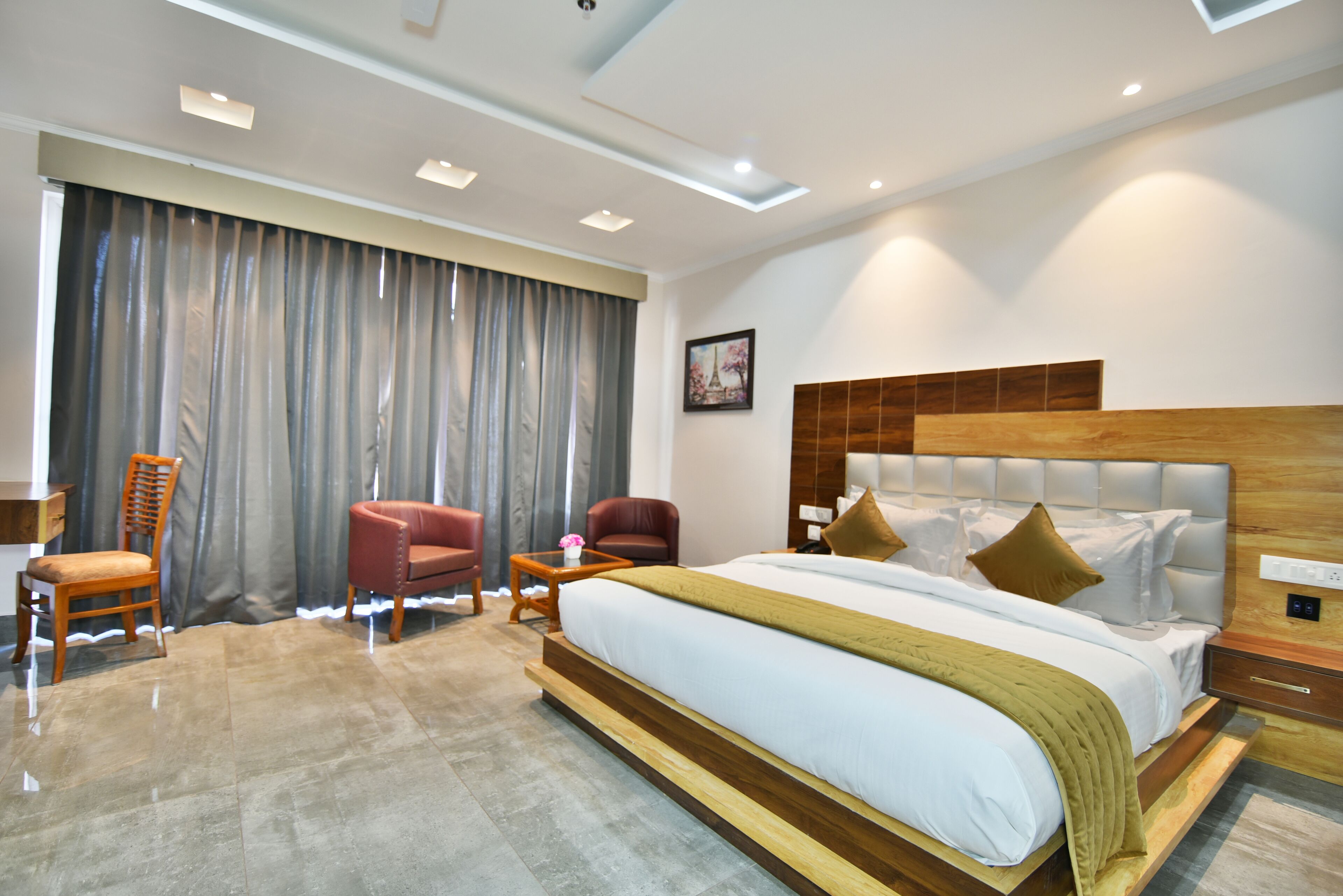 Quarto de hotel moderno com uma cama grande, duas poltronas, uma mesa pequena e uma janela ampla com cortinas.