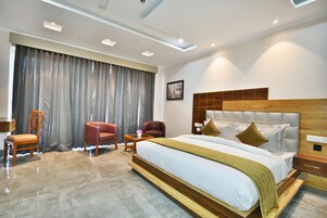Quarto de hotel moderno com uma cama grande, duas poltronas, uma mesa pequena e uma janela ampla com cortinas.