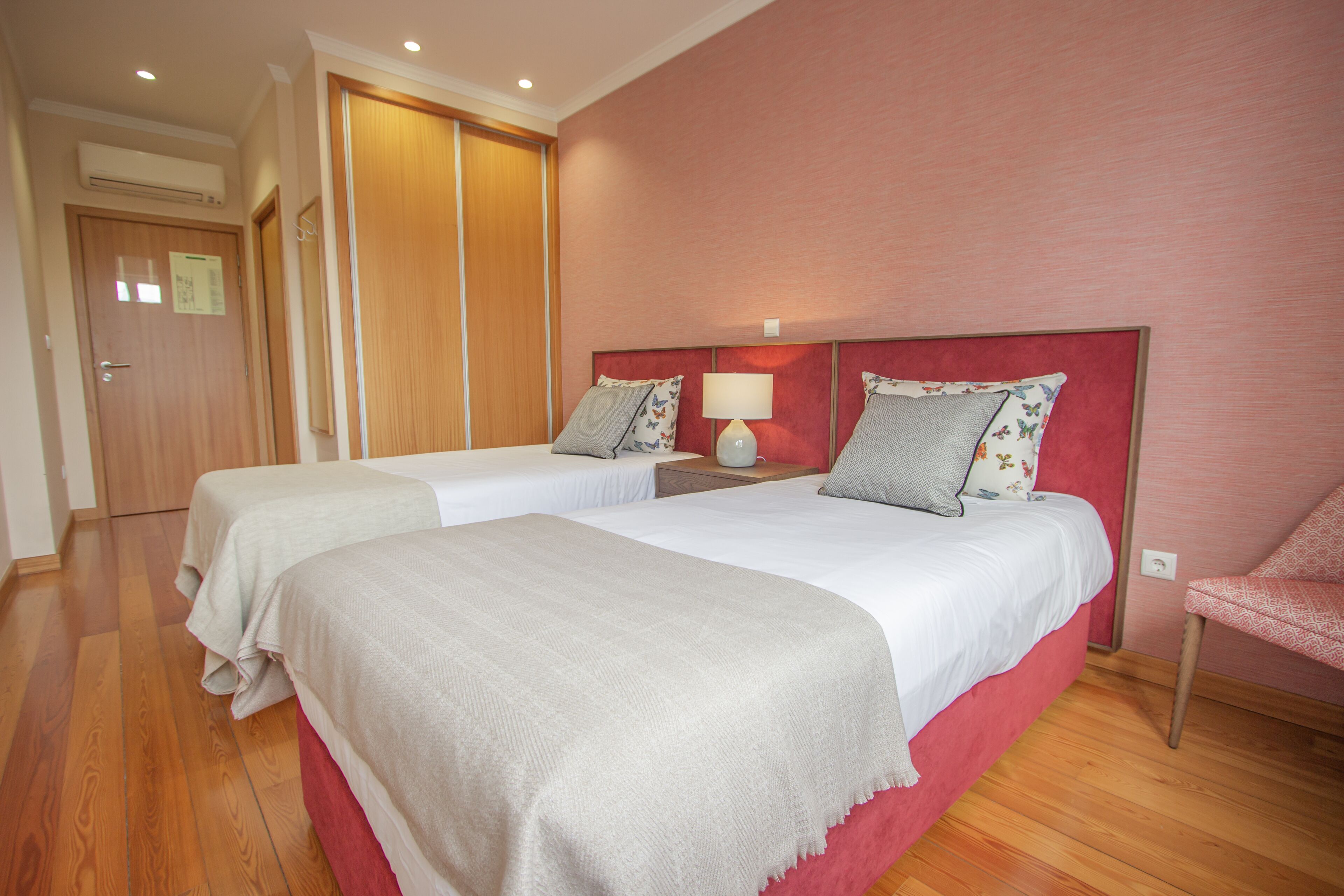 Quarto de hotel com duas camas, piso de madeira, parede de destaque vermelha e uma pequena mesa com abajur.