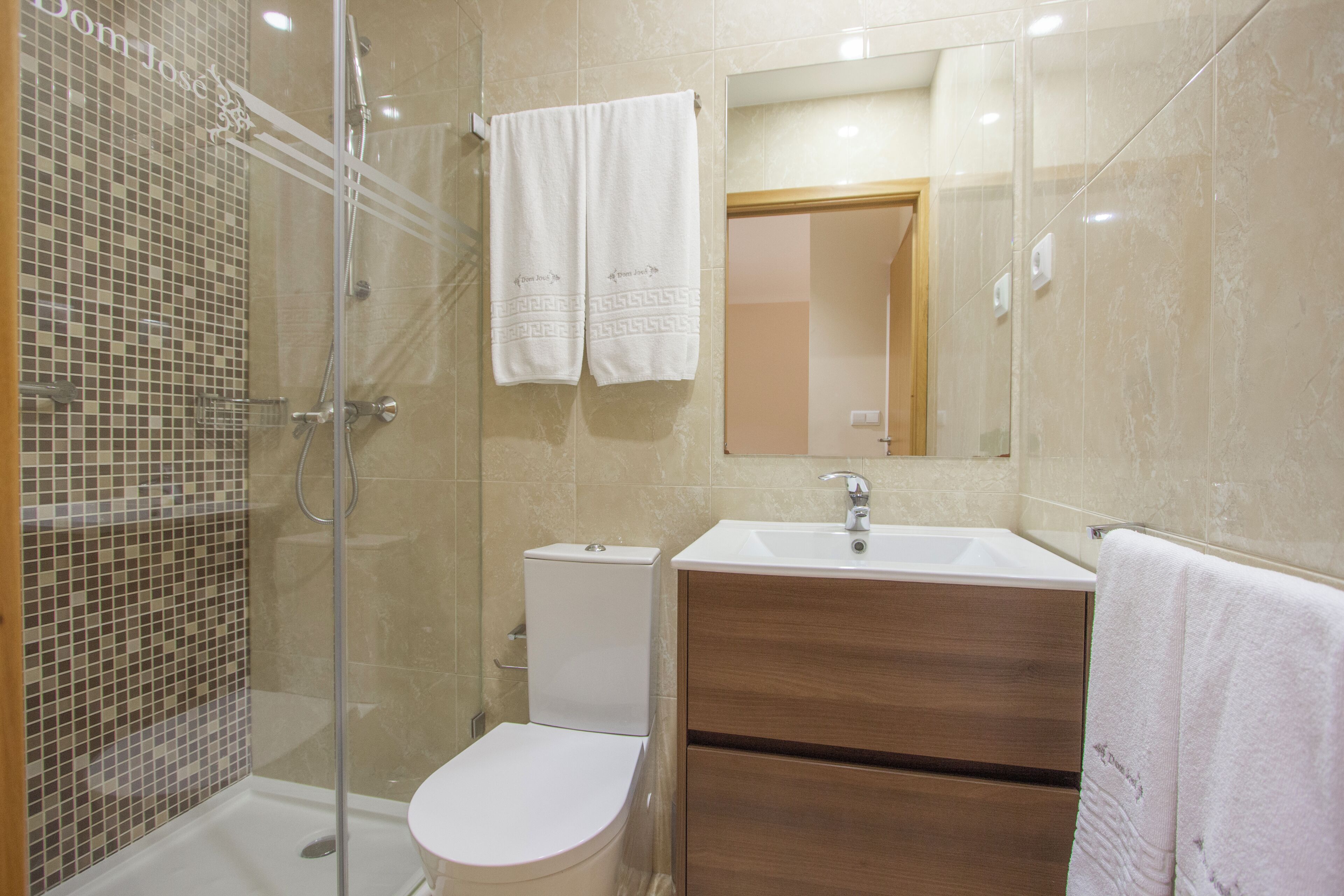 Uma casa de banho com chuveiro em cabine de vidro, sanita branca, toucador de madeira com espelho e duas toalhas brancas penduradas na parede.
