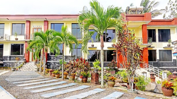 Um hotel com estrutura de dois andares, uma escadaria que leva à entrada e diversos vasos de plantas e palmeiras em primeiro plano.