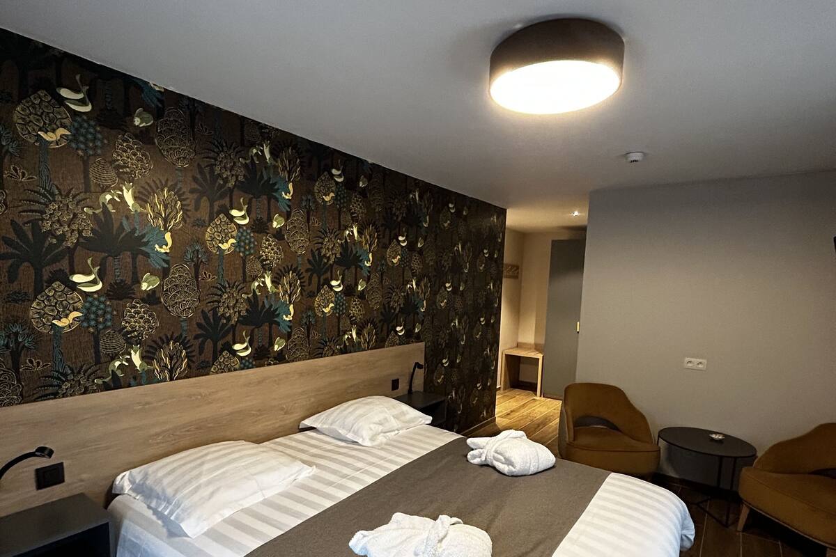 Quarto de hotel com uma cama grande, duas almofadas brancas, um cobertor cinza e duas toalhas brancas sobre a cama. Possui cabeceira de madeira, parede com um padrão decorativo e uma pequena mesa com uma cadeira.