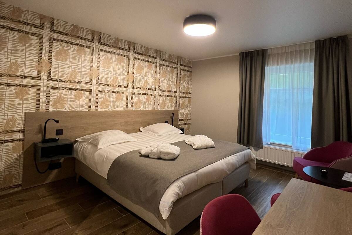 Quarto de hotel com piso de madeira, cama com roupa de cama branca, mesa de cabeceira com abajur, uma cadeira vermelha, uma mesa de madeira e uma janela com cortinas.