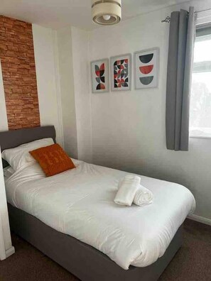 Uma cama bem arrumada com lençóis brancos e uma toalha dobrada, cabeceira cinza, parede de destaque em madeira e três obras de arte abstratas na parede.