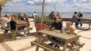 Uma área externa com mesas e bancos de madeira, plantas em vasos e vista para o mar.
