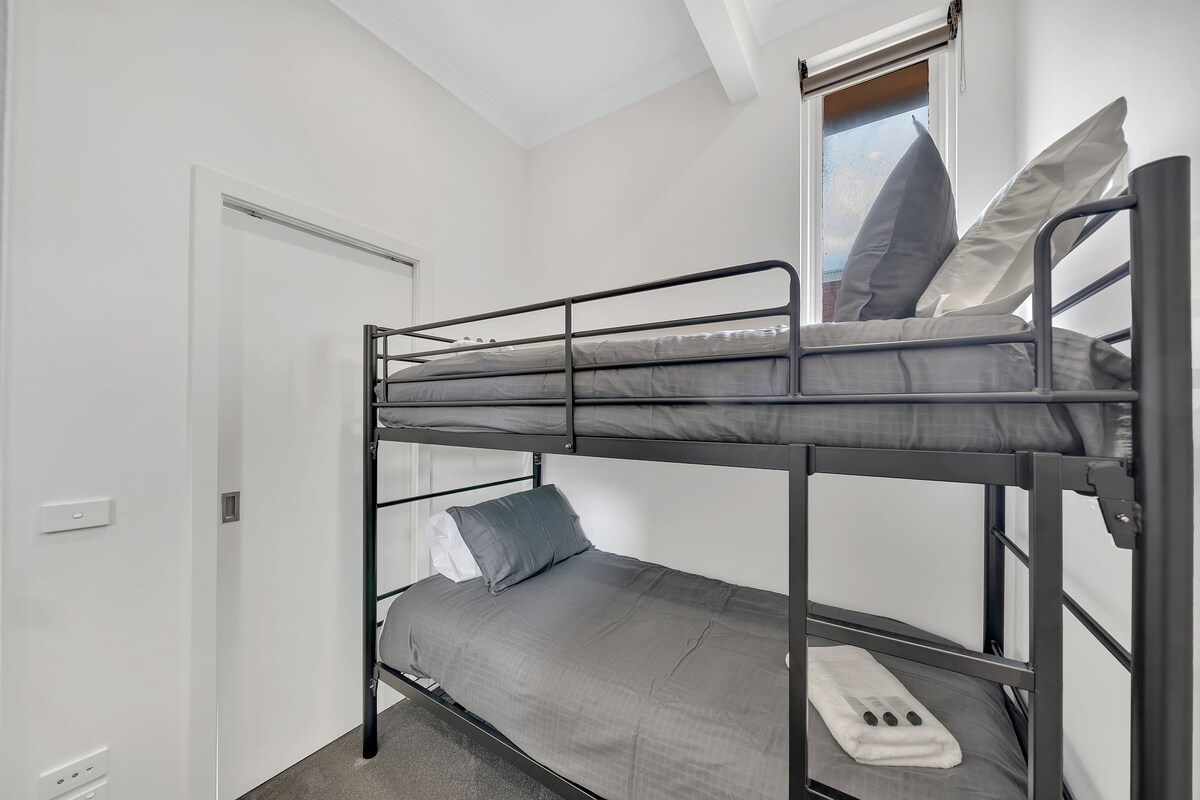 Uma cama beliche com roupa de cama e almofadas cinzas, uma porta branca e uma janela com persianas.