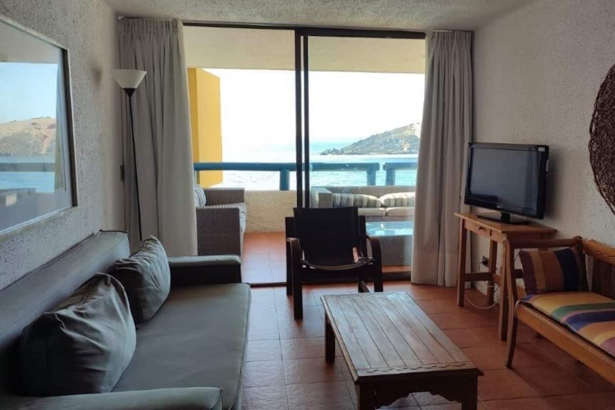Quarto com sofá, mesa de madeira, televisão e vista para o mar.