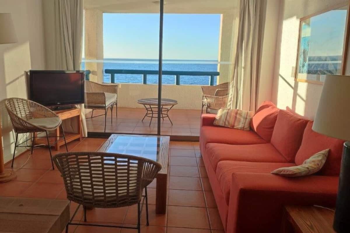 Quarto com sofá vermelho, cadeiras de vime, televisão e vista para o mar.
