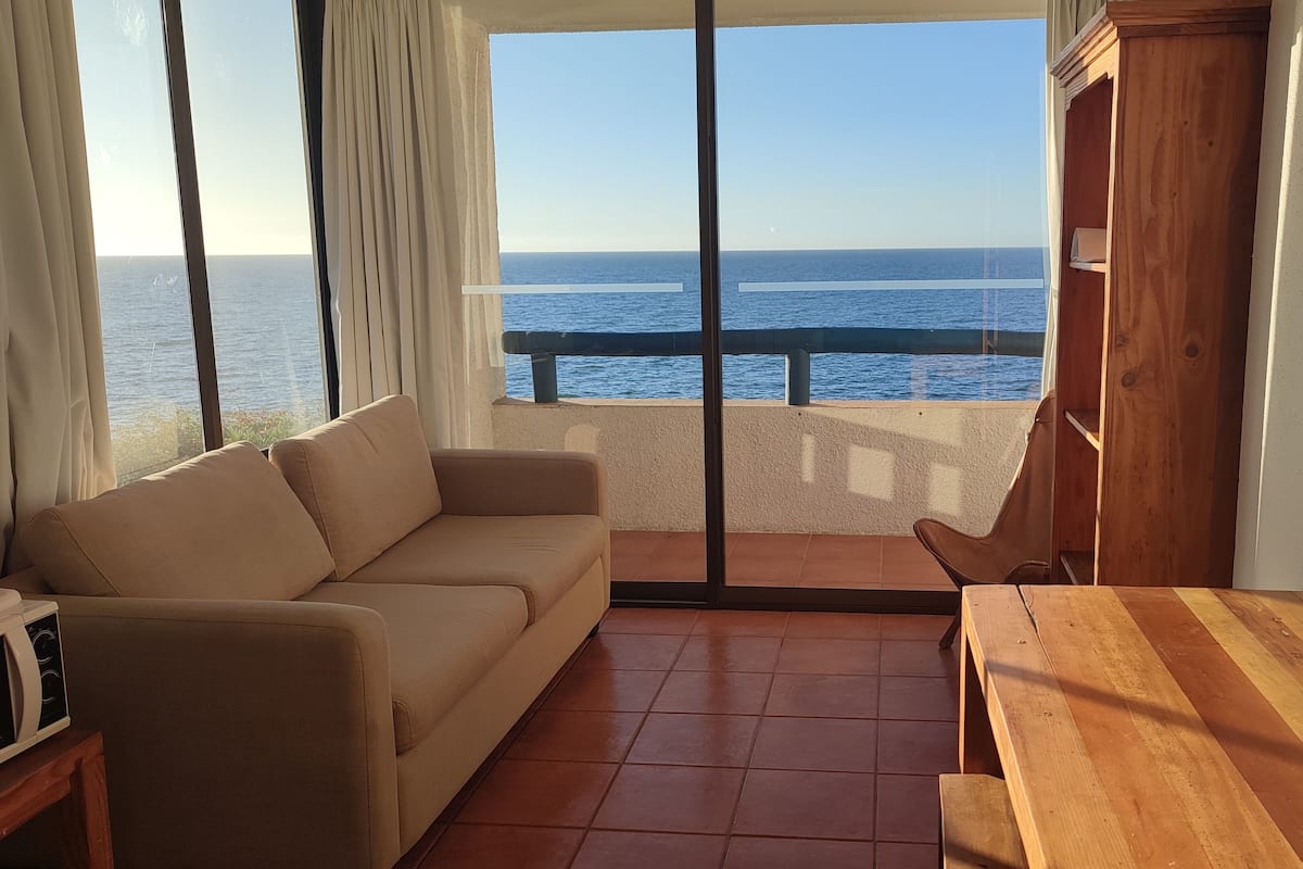 Quarto com sofá, mesa de madeira e vista para o mar através de amplas janelas.