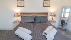 Uma cama bem arrumada com duas toalhas enroladas, duas luminárias de cabeceira e dois quadros na parede.