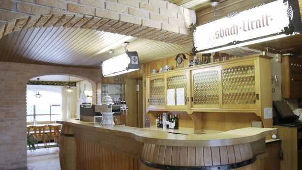 Uma área de bar com balcão de madeira, um letreiro acima com a inscrição "Liebach-Licht" e uma adega ao fundo.