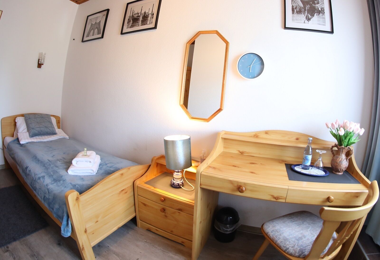 Um quarto pequeno e bem organizado, com uma cama, uma escrivaninha de madeira, uma cadeira, um espelho e um relógio na parede.