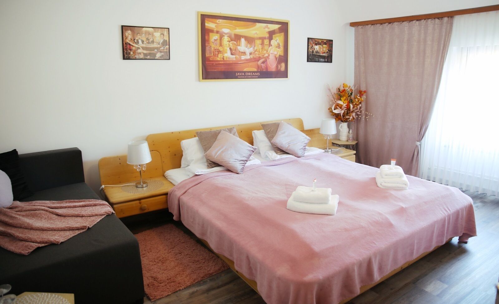 Quarto de hotel com cama, sofá, mesa de cabeceira e luminária. Há quadros na parede e uma janela com cortinas.