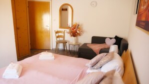 Quarto de hotel com cama, uma cadeira, uma mesinha com um vaso de flores e um relógio na parede.