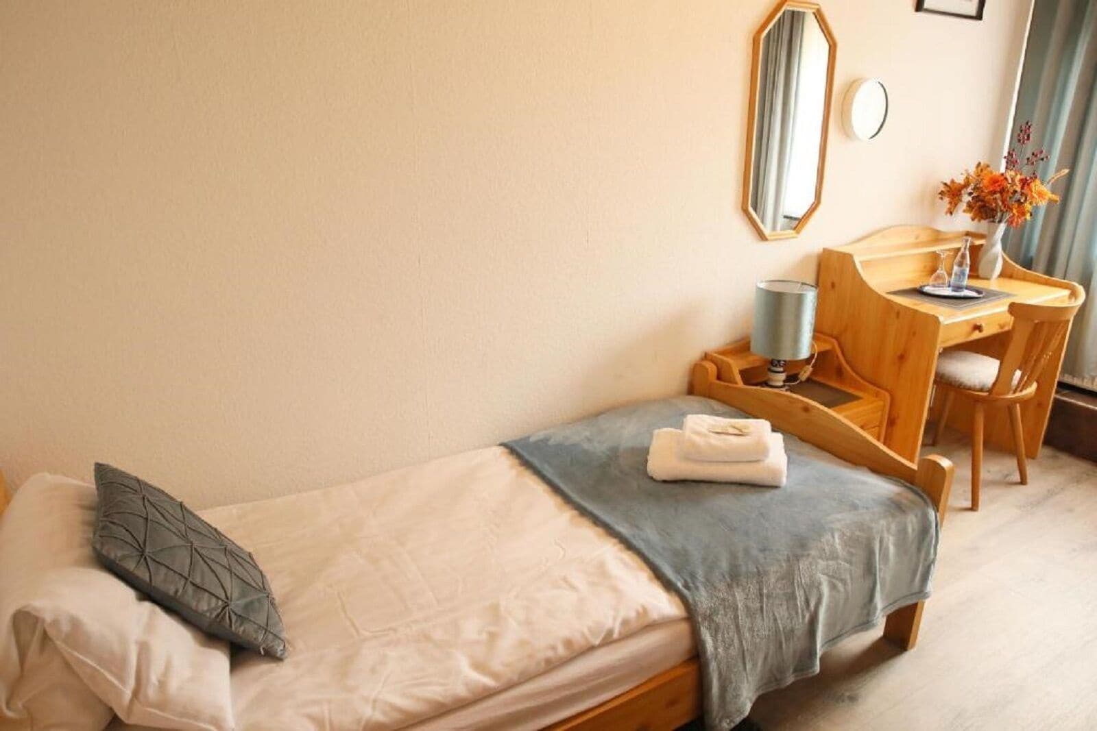 Cama bem arrumada com travesseiro cinzento e toalha dobrada, escrivaninha de madeira com cadeira, pequena mesa com um vaso de flores e espelho na parede.