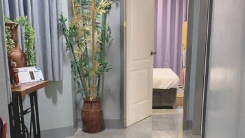 Um corredor com piso lustroso, uma mesa com computador, um vaso de planta e uma cama ao fundo.
