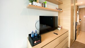 Quarto de hotel moderno com televisão de tela plana, estantes de madeira com livros e uma planta, minibar e placa de serviço de viagens.