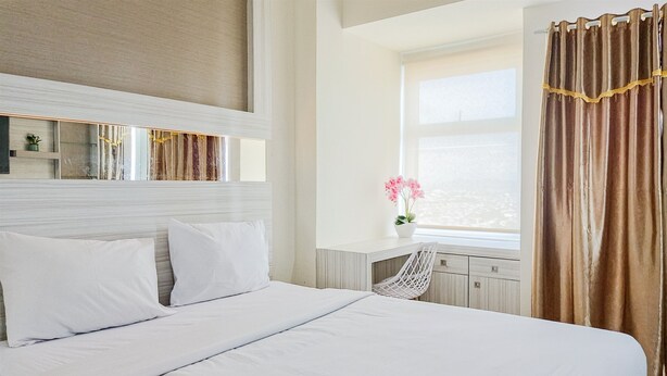 Uma cama bem arrumada com roupa branca, uma mesinha de cabeceira com um vaso de flores cor-de-rosa e uma janela com cortinas.