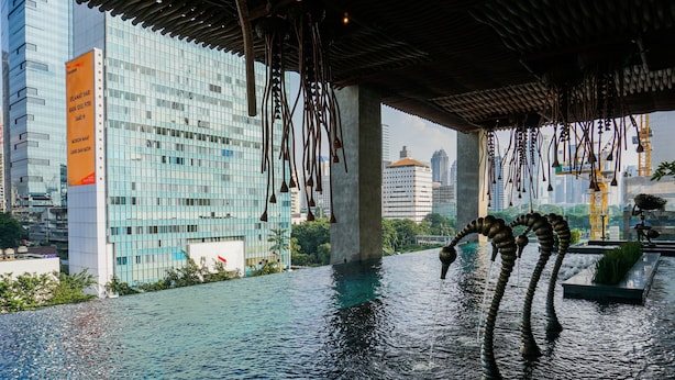 Um edifício moderno com fachada de vidro refletiva, um espelho d'água com uma escultura e vista para a paisagem urbana.