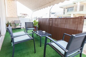 Um pátio externo com mesa e cadeiras, área coberta e vista para prédios ao fundo.