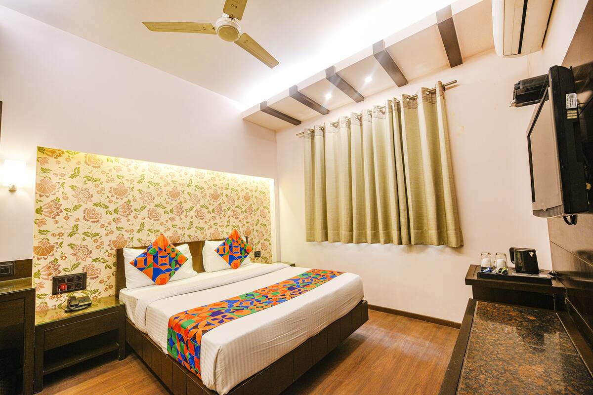 Quarto de hotel com cama, mesa de cabeceira, ventoinha no teto, janela com cortinas e televisão na parede.