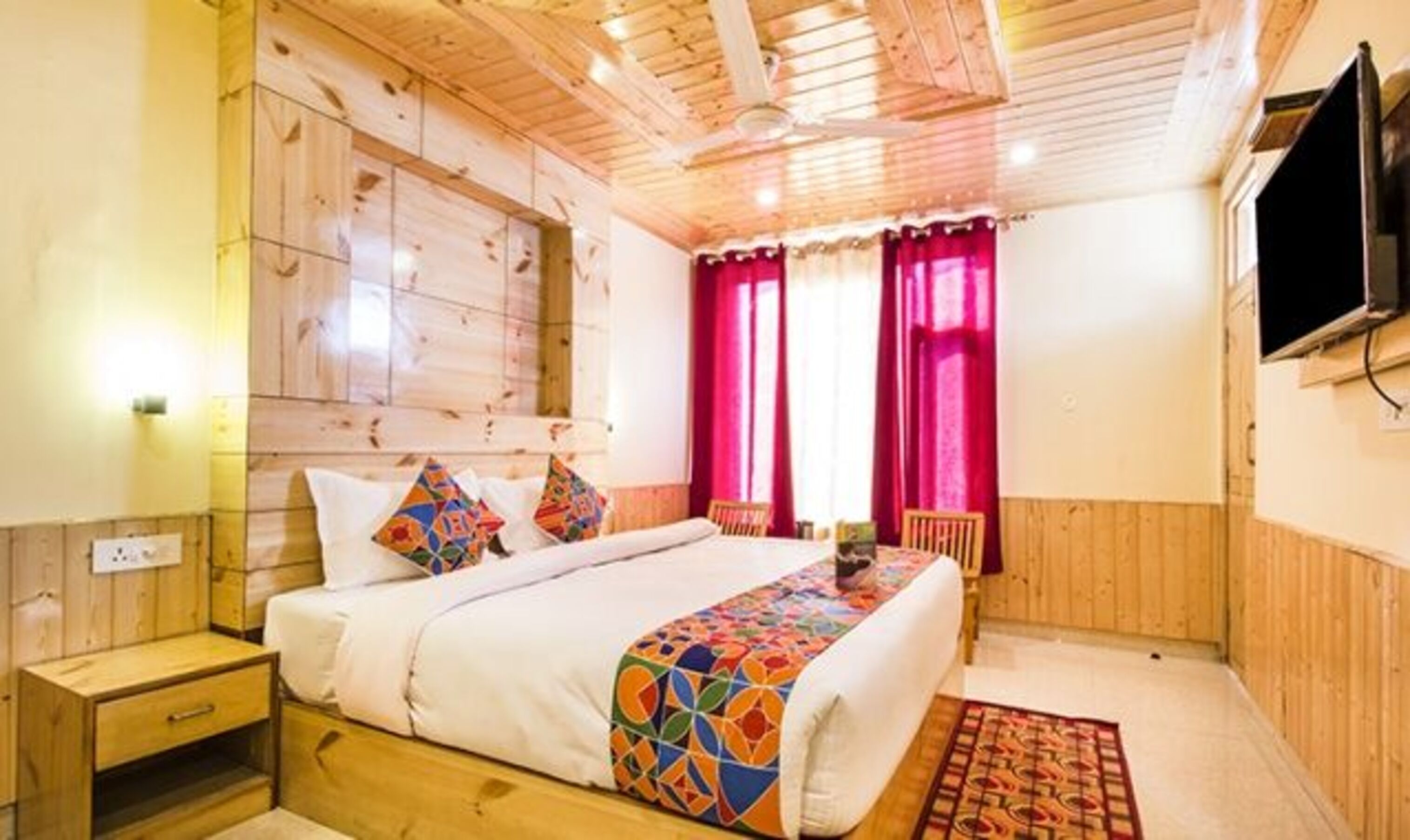 Quarto com painéis de madeira, uma cama, mesa de cabeceira, televisão e janela com cortinas.