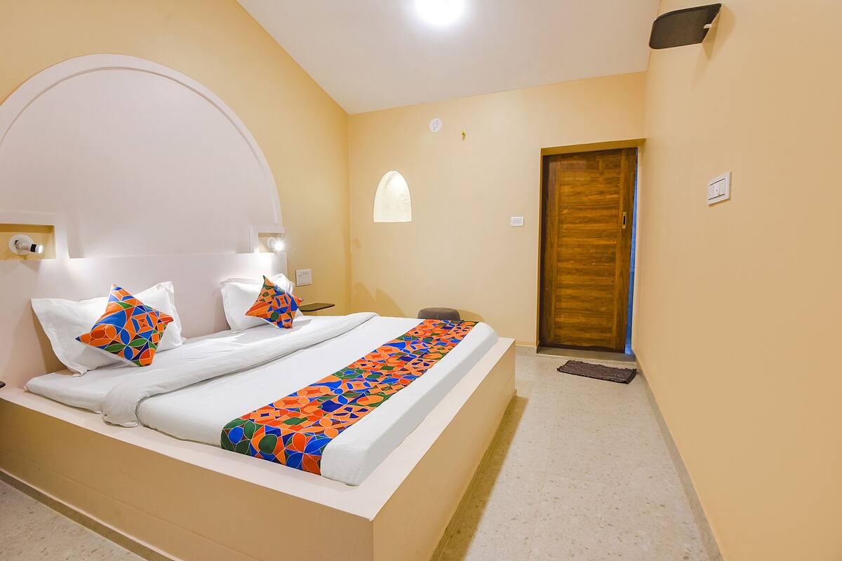 Quarto de hotel com cama, porta de madeira, janela pequena e luminária fixada na parede.
