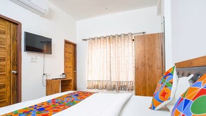 Quarto de hotel com uma cama, televisão na parede, guarda-roupa de madeira, janela com cortinas e mesa de cabeceira.