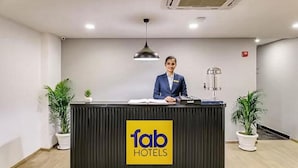 Uma mesa de recepção com o logotipo da Fab Hotels, duas plantas em vasos, uma luminária pendente moderna e um objeto de vidro transparente no balcão.