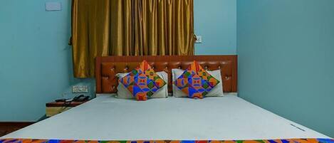 Quarto com cama, duas almofadas estampadas com padrões geométricos, janela com cortinas e ar condicionado fixado na parede.