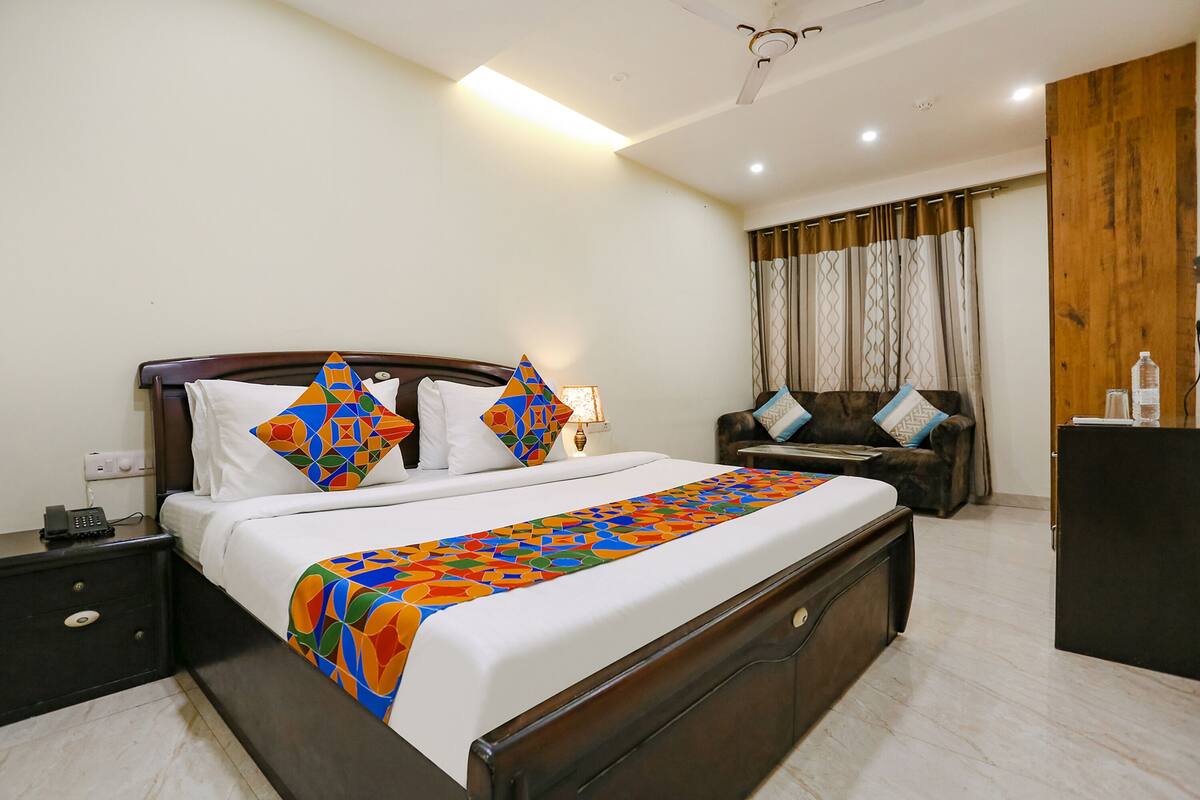 Quarto de hotel com uma cama grande, mesa de cabeceira com telefone, criado-mudo, encosto de cama de madeira, escrivaninha com abajur e uma cadeira.