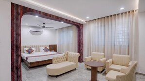 Quarto de hotel moderno com cama, sofá, duas poltronas, mesa redonda e ventilador de teto.