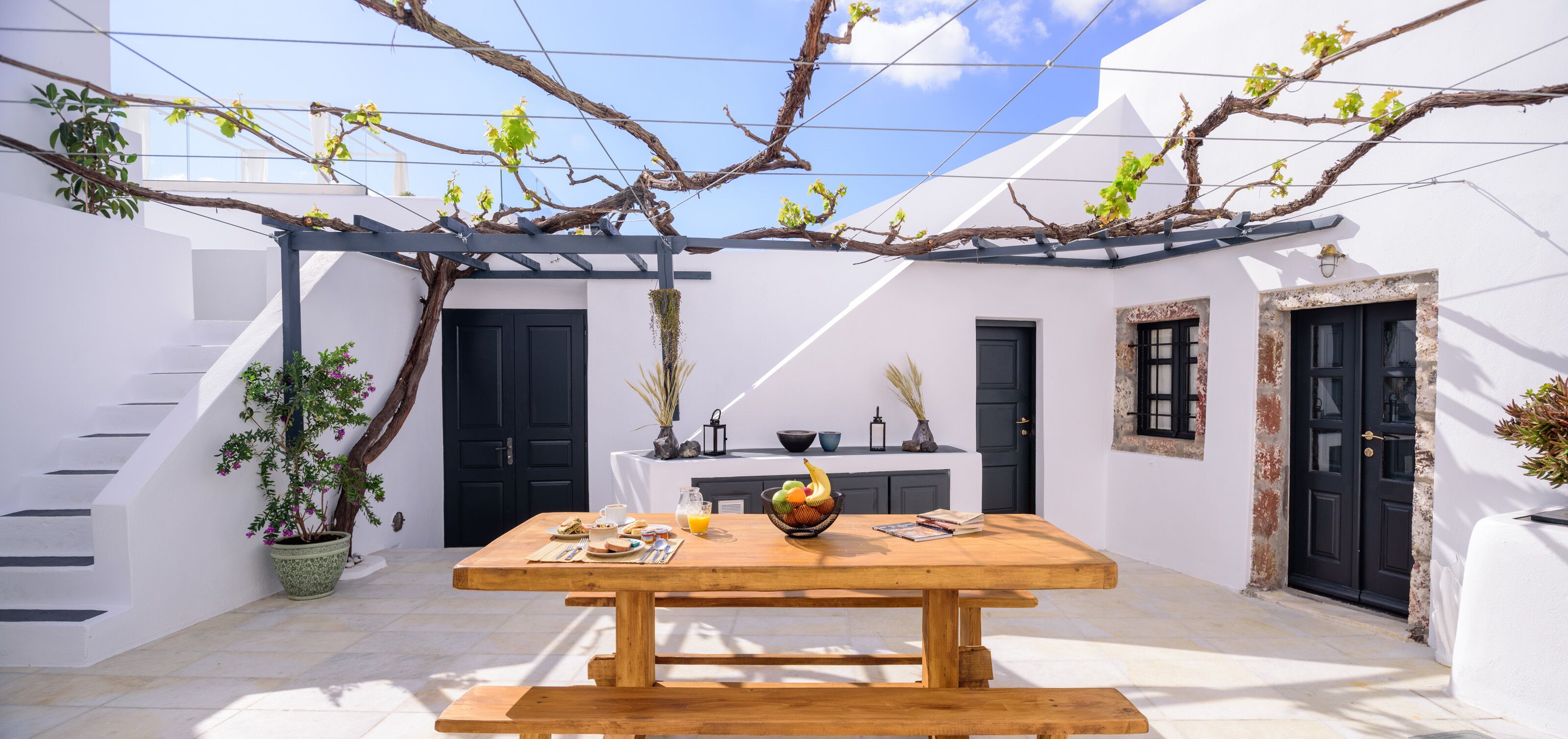 Um pátio com uma mesa de madeira posta para o café da manhã, cercado por paredes brancas e portas pretas.