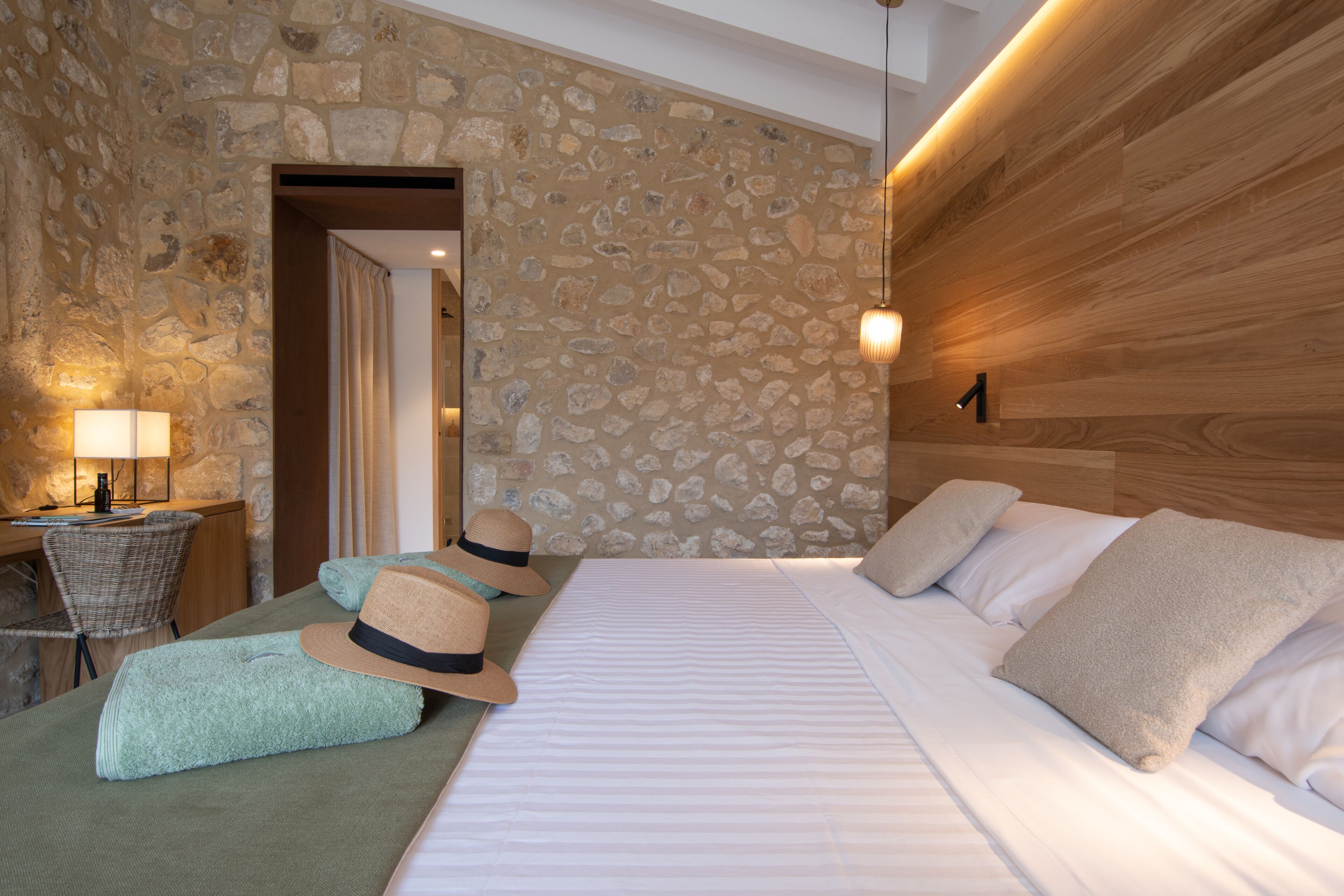 Quarto de hotel com parede de pedra, cabeceira de madeira, cama com roupa branca, escrivaninha com abajur e cadeira de vime.