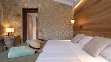 Quarto de hotel com parede de pedra, cabeceira de madeira, cama com roupa branca, escrivaninha com abajur e cadeira de vime.
