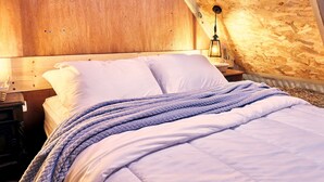 Uma cama com lençóis brancos e manta de tricô azul, uma cabeceira de madeira e uma mesa de cabeceira com abajur.