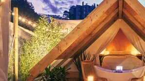 Uma tenda de madeira aconchegante em formato triangular, com um espaço de estar acolchoado e uma vela acesa, diante de um céu crepuscular.