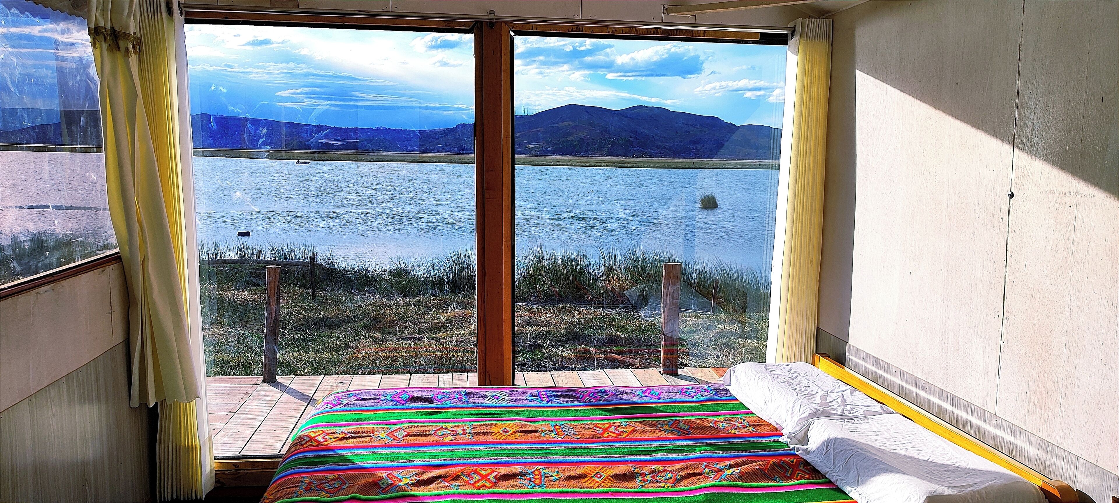 Quarto com uma grande janela com vista para um lago e montanhas, uma cama com um cobertor colorido, e um deck de madeira na parte externa.