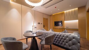 Quarto de hotel moderno com sofá estofado, mesa de jantar com cadeiras, televisão de tela plana na parede e piso de madeira.