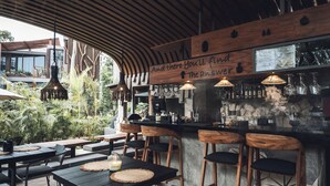 Uma área de bar ao ar livre com móveis de madeira, parede de pedra e luminárias pendentes.