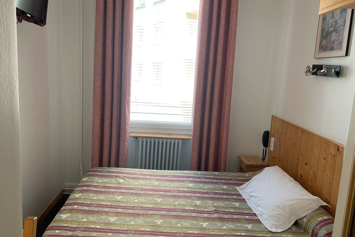 Um quarto com cama, mesa de cabeceira, abajur, janela com cortinas e radiador.